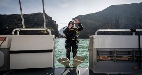 Girl on a boat in scruba diving gear falling backwards with Carrick-a-Rede Rope Bridge in the background as part of a scuba diving experience with Aqu
